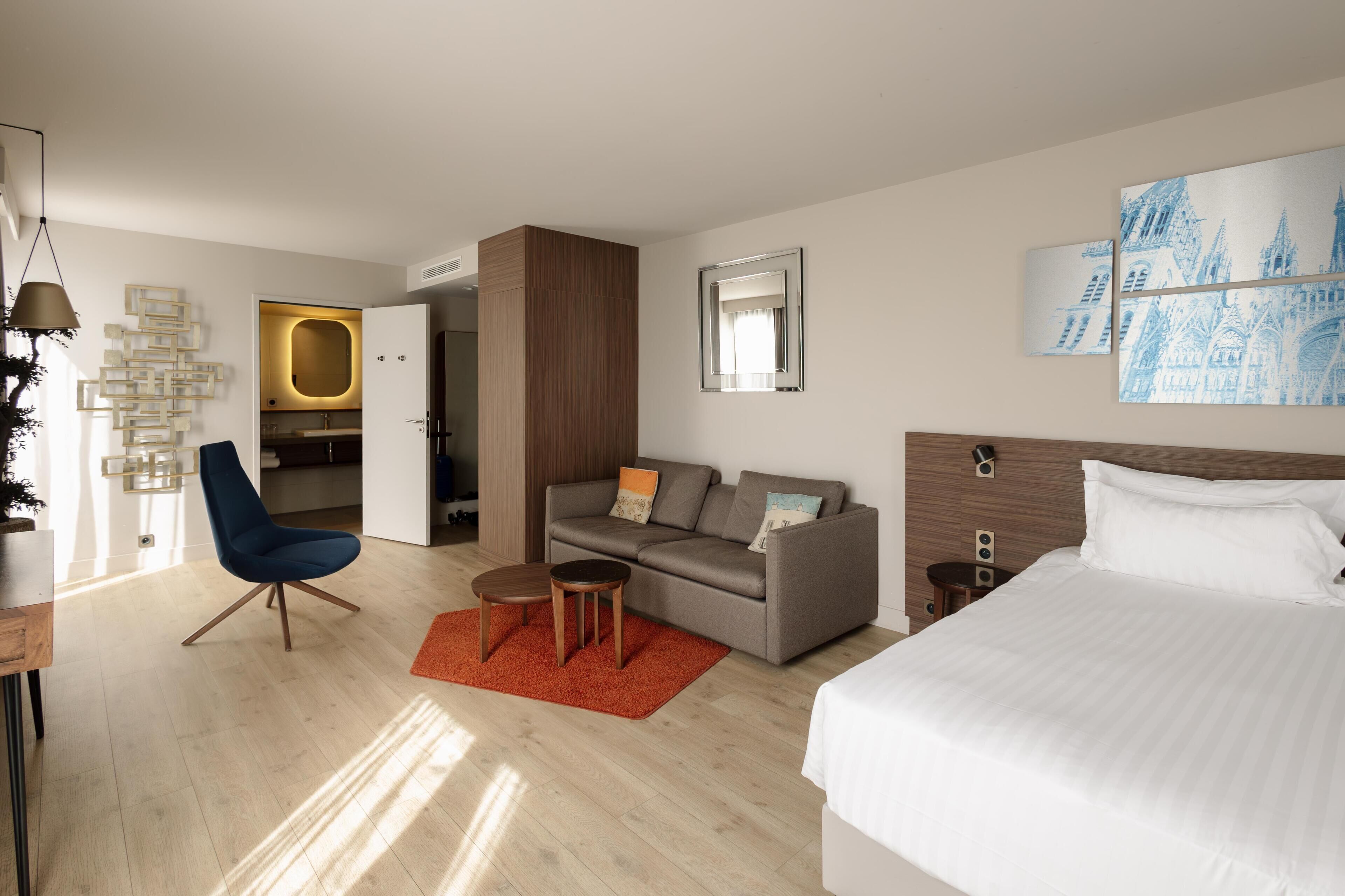 Quarto de hotel moderno com uma cama, sofá, cadeira, mesa pequena e um quadro na parede.
