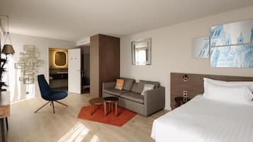 Quarto de hotel moderno com uma cama, sofá, cadeira, mesa pequena e um quadro na parede.