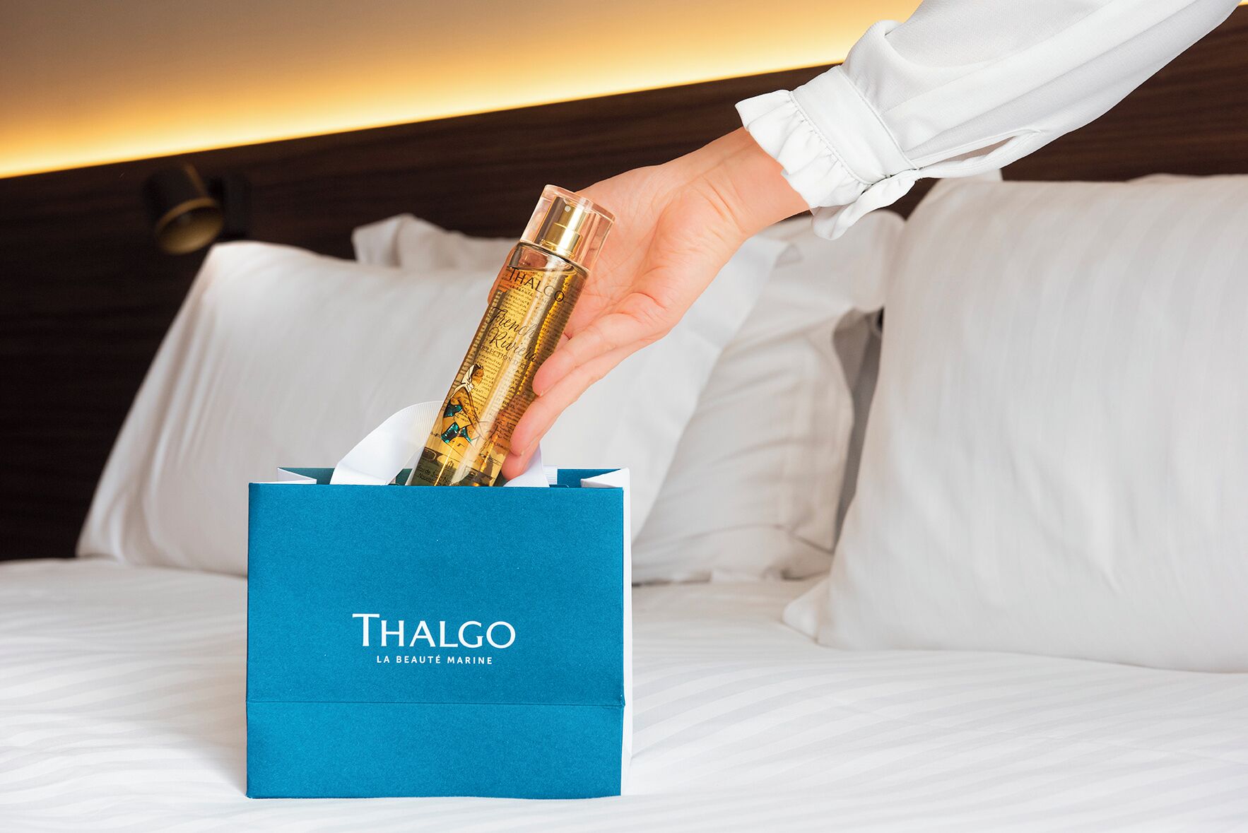 Uma mão colocando uma garrafa em uma sacola azul Thalgo sobre uma cama com almofadas brancas e cabeceira escura.