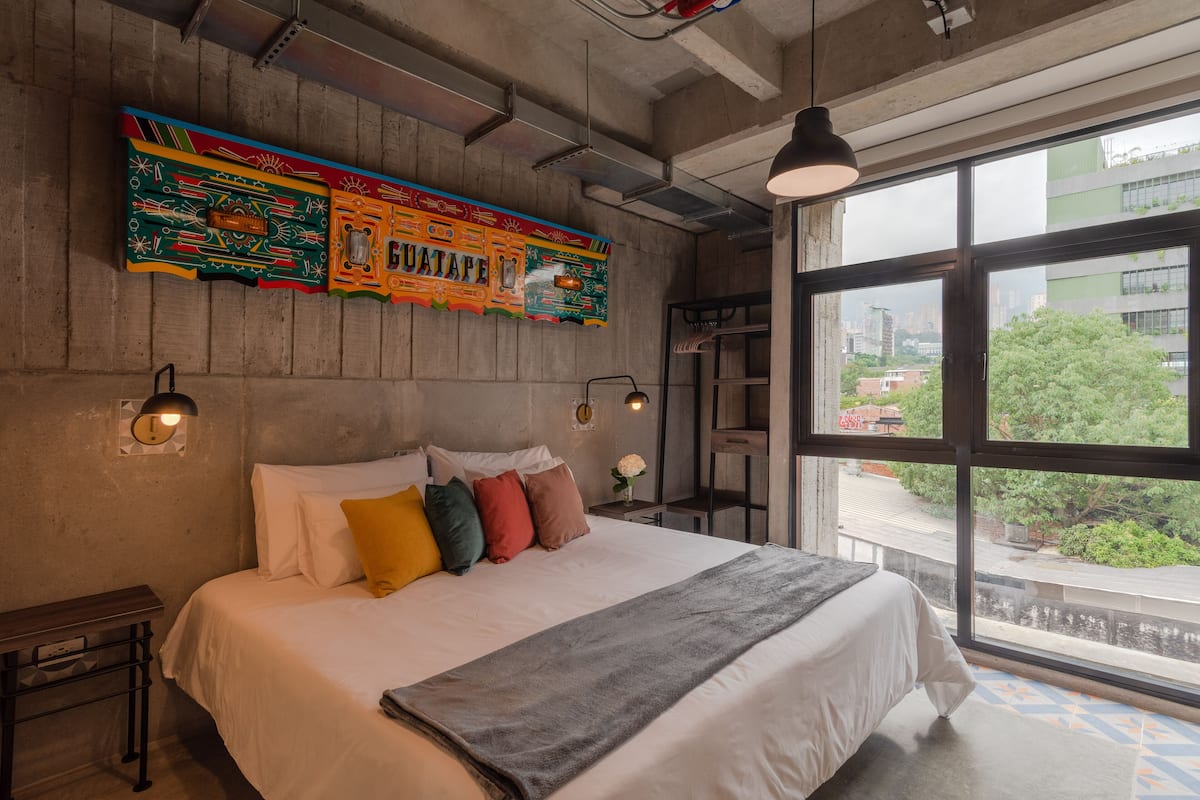 Quarto com uma cama grande, uma decoração de parede colorida, uma janela com vista para edifícios e árvores, e uma mesinha com abajur.