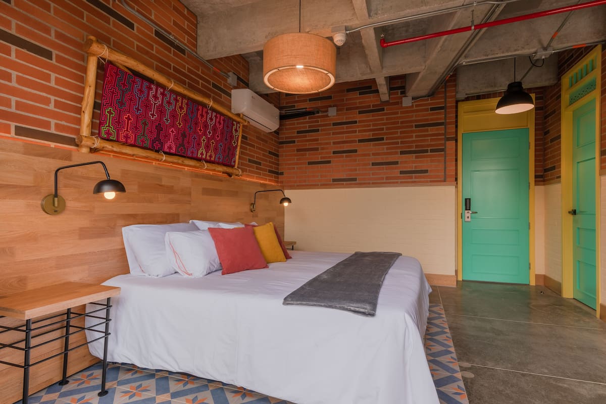 Quarto com uma cama, uma mesinha de cabeceira de madeira, parede estampada, luminária pendente e uma porta verde.