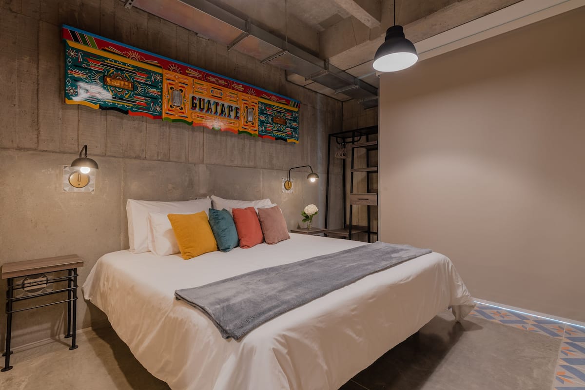 Uma cama com roupa de cama branca, almofadas coloridas e um cobertor cinza. Duas luminárias fixadas na parede estão de cada lado da cama. Uma tapeçaria colorida está pendurada acima da cama.