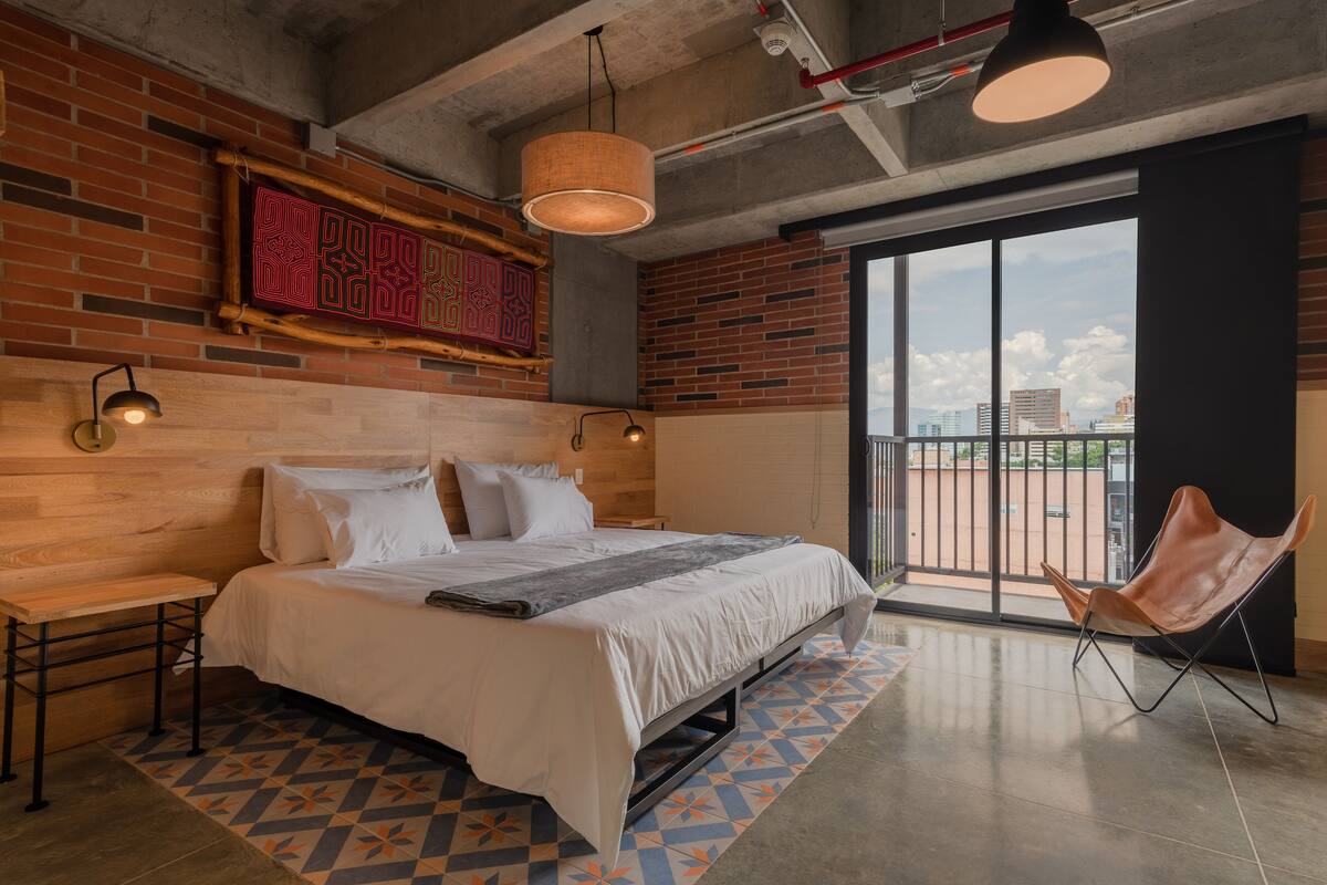Quarto de hotel moderno com uma cama grande, cabeceira de madeira, tapete estampado e vista da cidade pela varanda.