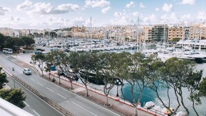 Uma marina com vários barcos atracados, uma estrada com veículos e edifícios ao fundo.