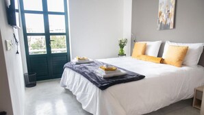 Quarto de hotel moderno com uma cama grande, roupa de cama branca, um cobertor escuro e uma pequena mesa de cabeceira.