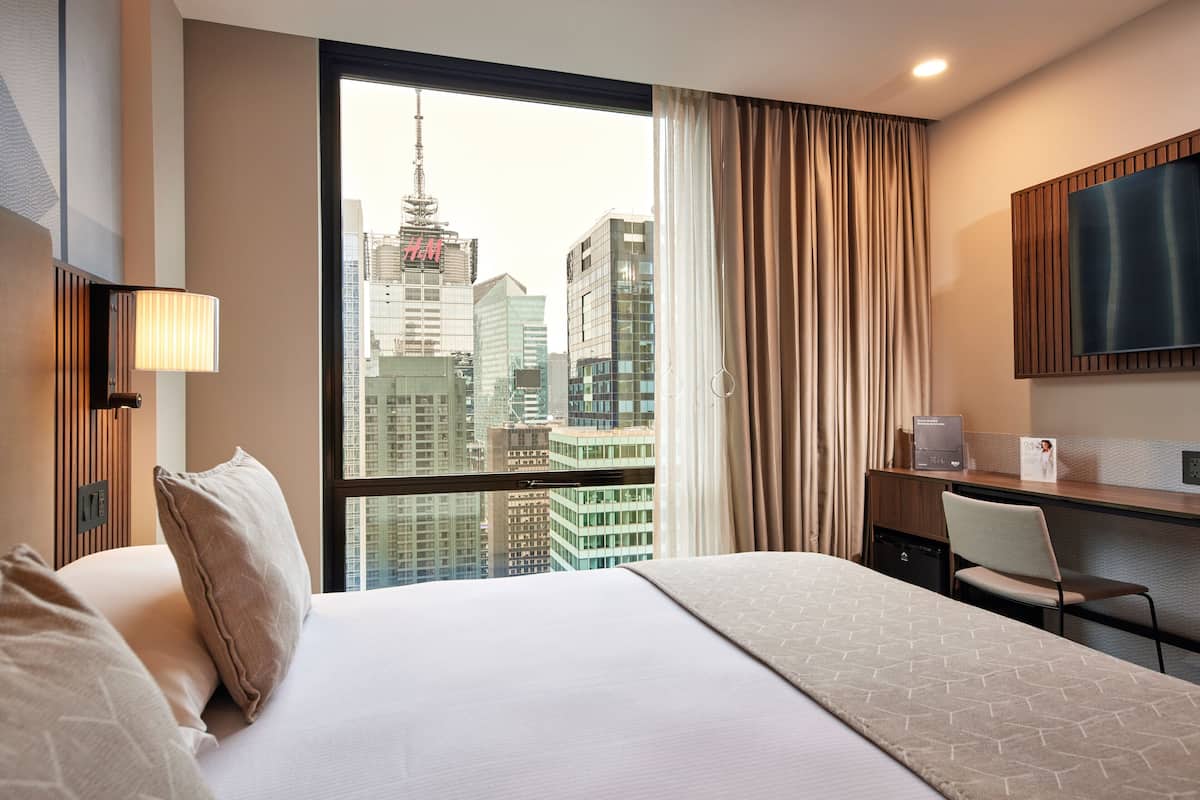 Quarto de hotel com uma cama grande, uma escrivaninha, uma cadeira, uma televisão e vista para a paisagem urbana.