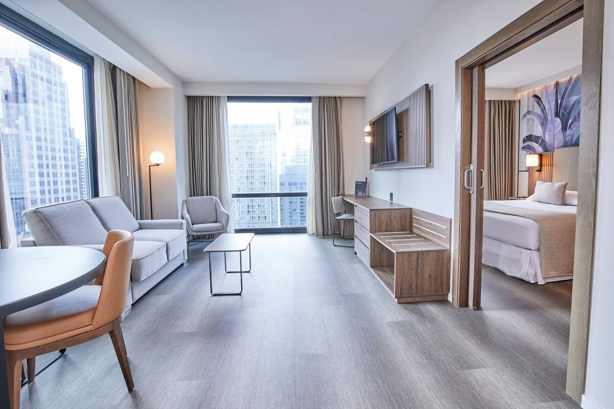 Quarto de hotel moderno com sofá, mesa de jantar, escrivaninha, cama e uma grande janela com vista para a cidade.