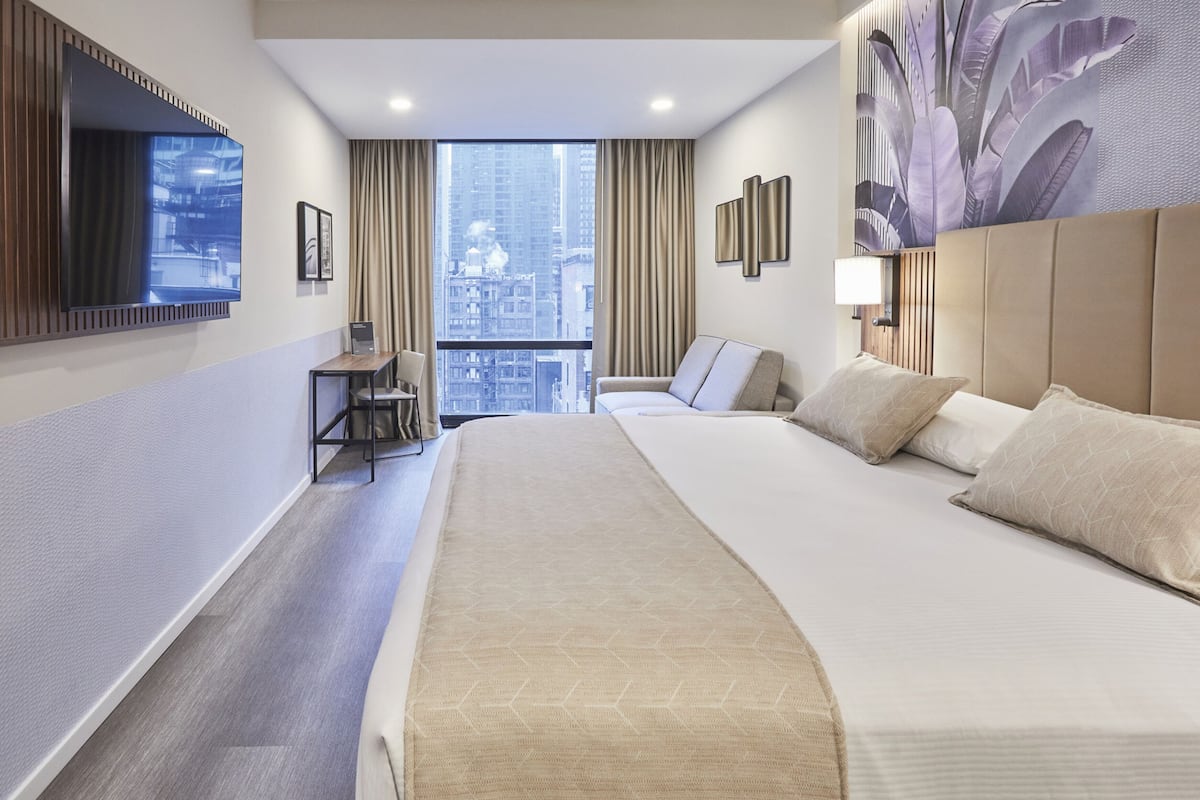 Quarto de hotel com uma cama grande, uma escrivaninha, uma cadeira e uma janela ampla com vista para a cidade.