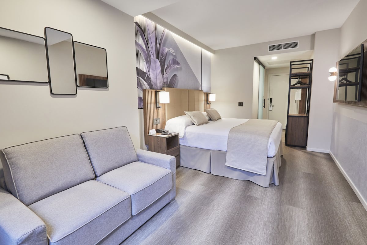 Quarto de hotel moderno com sofá, cama, mesa de cabeceira e um espelho grande.