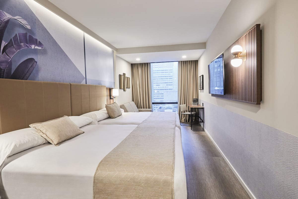 Quarto de hotel com duas camas, televisão, escrivaninha e janela grande com cortinas.