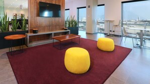 Um hall de hotel moderno com tapete vermelho, pufes amarelos, um banco de madeira e uma TV de tela plana na parede.