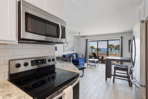 Cozinha moderna com micro-ondas e fogão embutidos, sala de jantar com mesa e cadeiras, e varanda com vista para a praia.