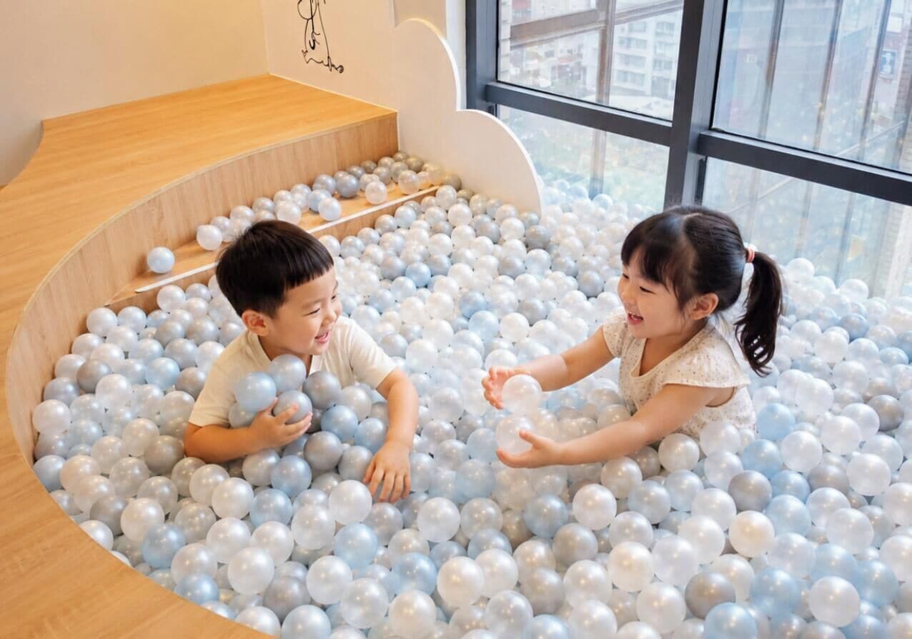 Duas crianças brincam em uma piscina de bolinhas, cheia de bolinhas brancas e azul claro, com um banco de madeira e uma janela grande ao fundo.