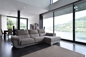 Uma sala de estar moderna com um amplo sofá em forma de L, uma área de refeições com mesa e cadeiras, e grandes janelas com vista para um lago e colinas.
