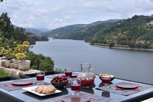 Disposição para refeições ao ar livre com vista panorâmica para um lago e colinas, incluindo uma mesa com pratos, copos, um jarro e uma tigela de cerejas.