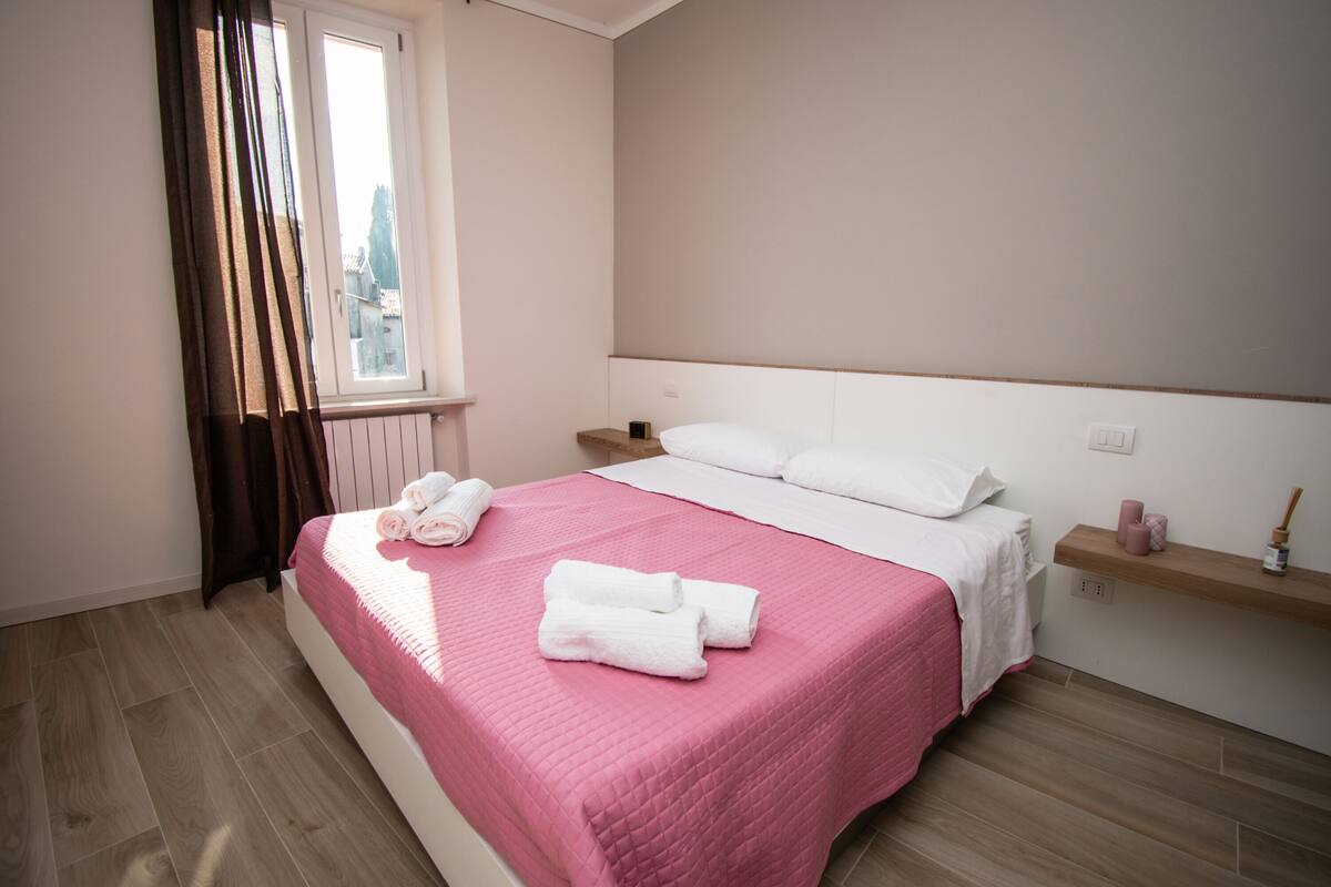 Uma cama bem arrumada com roupa de cama rosa e toalhas brancas, piso de madeira, uma janela com cortinas e uma pequena prateleira com produtos de higiene pessoal.