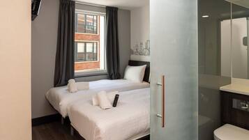 Quarto de hotel com duas camas, uma janela grande com cortinas, banheiro com box de vidro e um mural na parede retratando uma paisagem urbana.