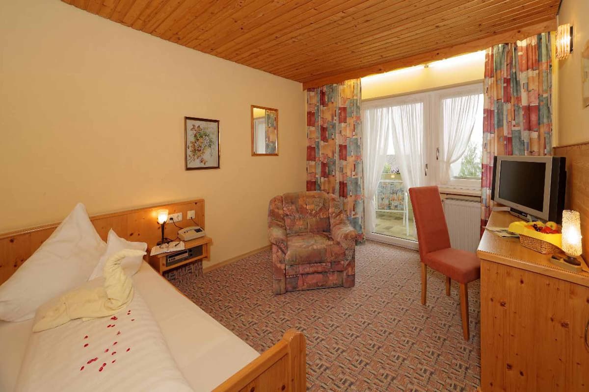 Quarto de hotel com cama, cadeira, escrivaninha com televisão e janela com cortinas.
