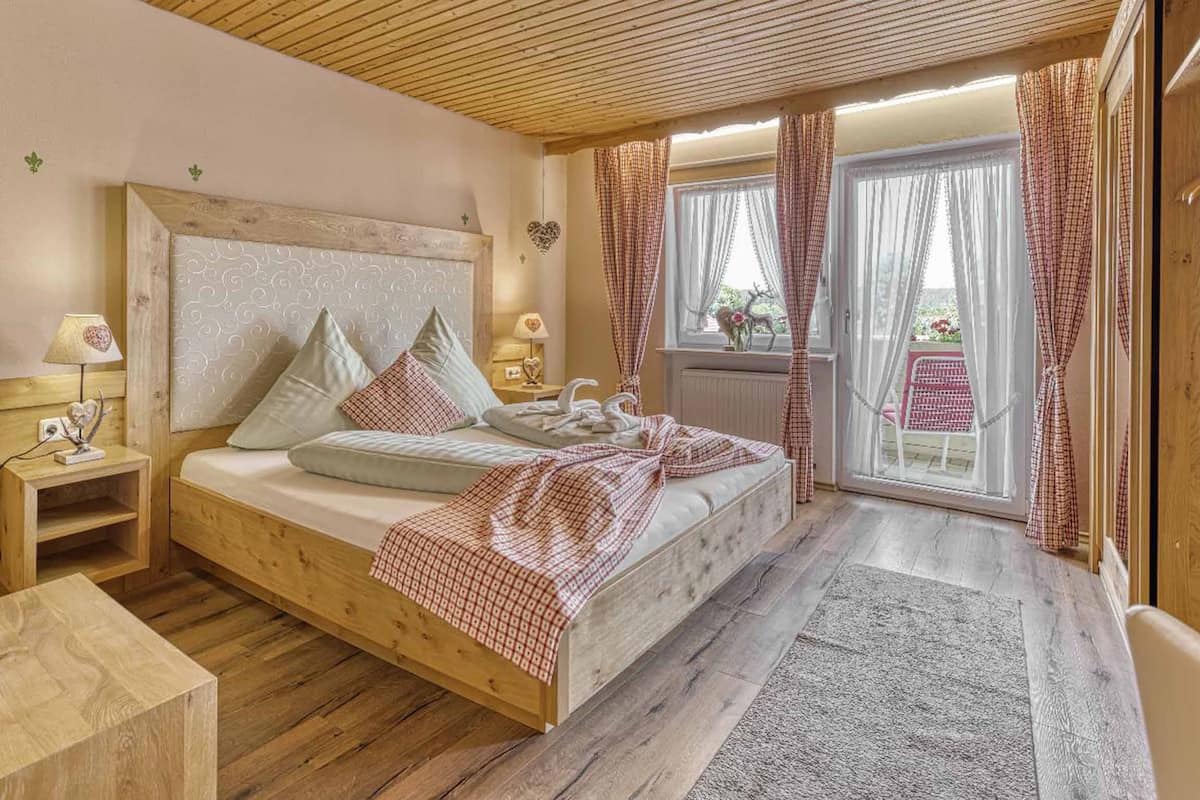 Um quarto aconchegante com cabeceira de madeira, cama com cobertor xadrez, mesa de cabeceira com luminária e vista para uma varanda com cortinas.