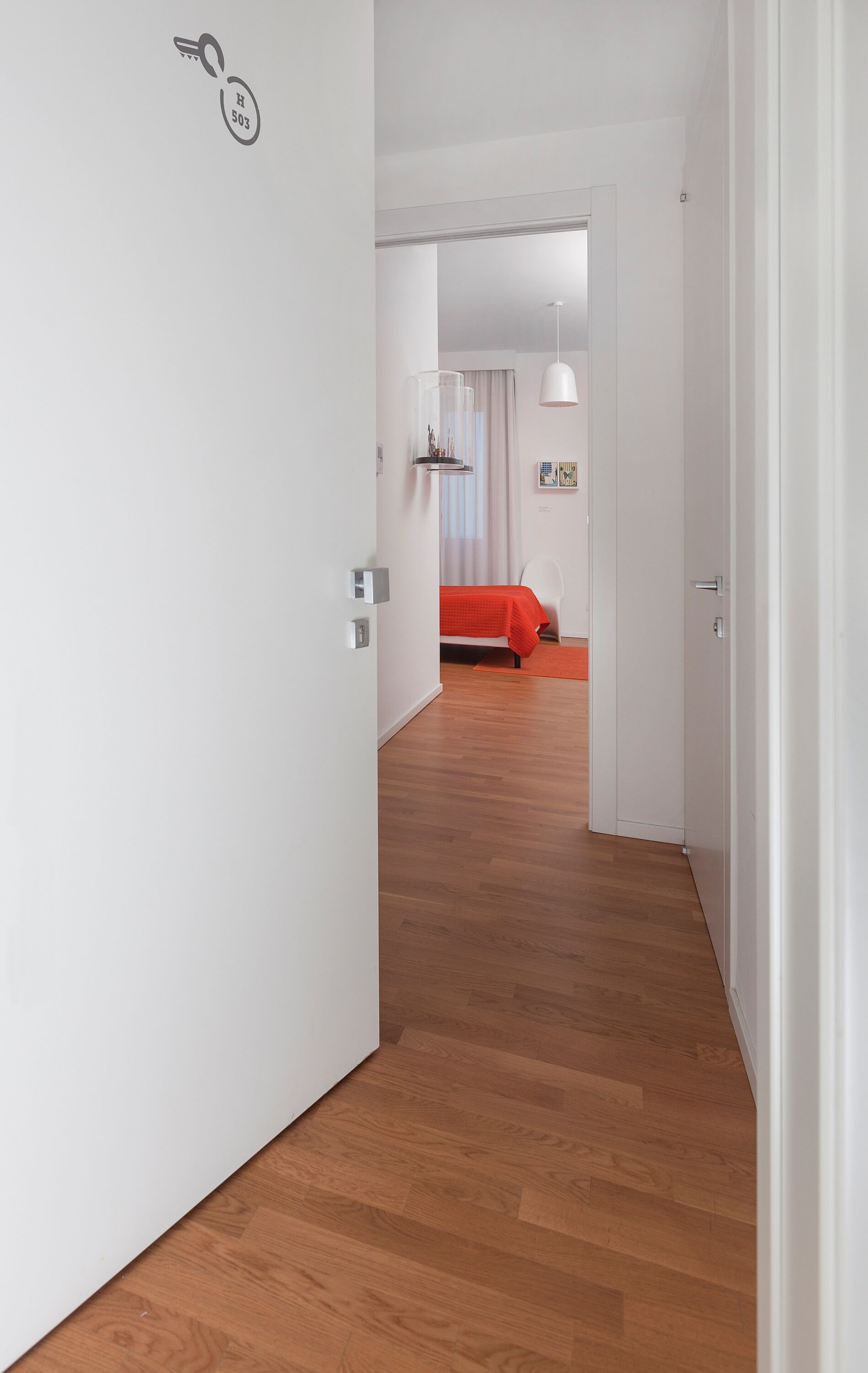 Quarto de hotel moderno com piso de madeira, cama com colcha vermelha, luminária pendente e prateleira na parede com artigos de higiene pessoal.