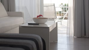 Quarto de hotel moderno com sofá branco, uma mesa pequena com uma tigela e um controle remoto, e uma varanda com corrimão e plantas em vasos.