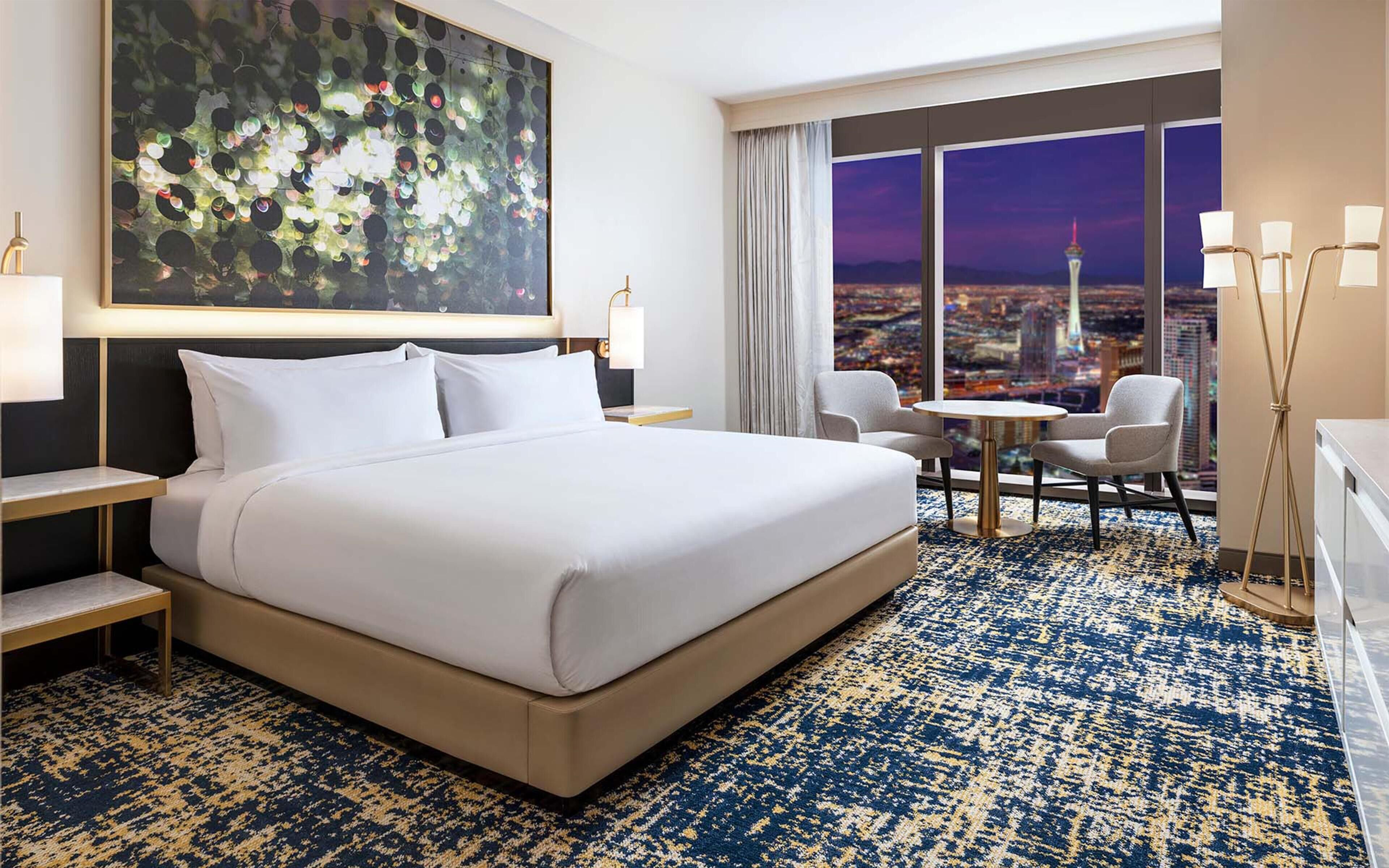 Quarto de hotel com cama grande, escrivaninha, cadeira e vista para o horizonte urbano ao entardecer.