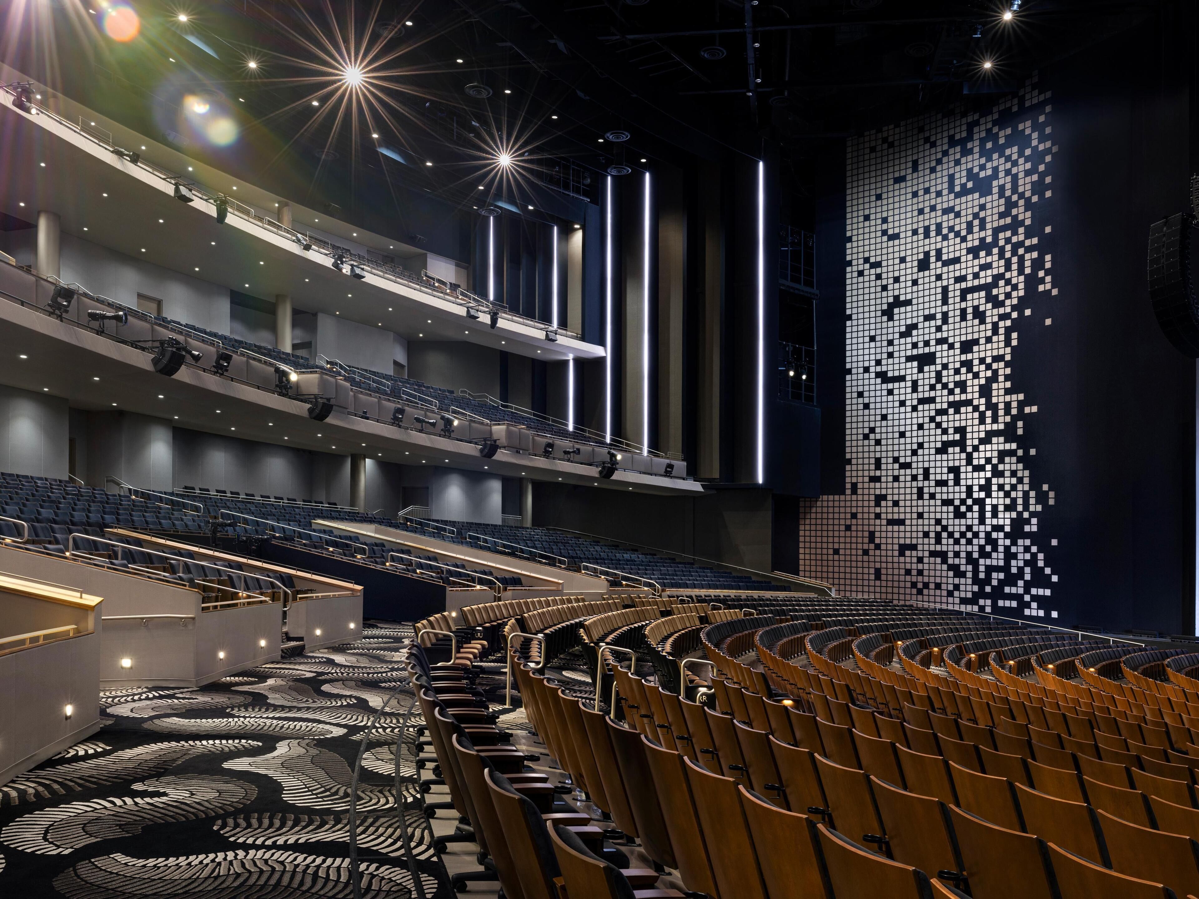 Um teatro moderno com assentos em níveis, um carpete estampado e uma grande tela digital exibindo um design pixelizado.