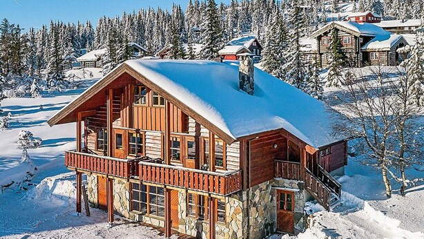 Uma cabana de tronco com varanda, cercada por árvores cobertas de neve e um céu claro.