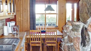 Interior de cabana de madeira com parede de pedra, mesa de jantar com cadeiras, luminária pendente e janela com vista para árvores.