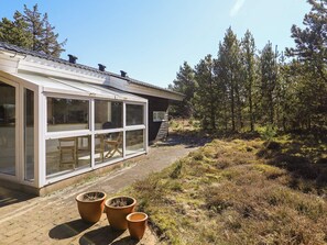 Uma casa moderna com grandes janelas de vidro, área de pátio com plantas em vasos e paisagem arborizada.