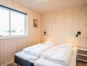 Quarto com paredes de madeira, cama com roupa de cama branca, duas luminárias laterais e janela com vista para edifícios.