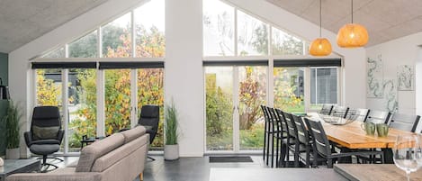 Quarto amplo com grande mesa de jantar, cadeiras modernas, sofá e janelas amplas com vista para um jardim.