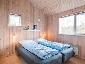 Quarto com paredes de madeira, cama com roupa de cama azul, duas luminárias na parede, uma janela e uma prateleira com objetos decorativos.