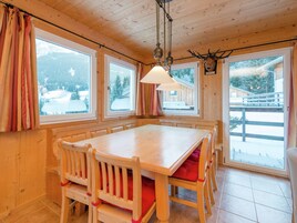Uma sala de jantar de madeira com uma mesa grande e cadeiras, janelas amplas com cortinas e um teto com luminárias pendentes.