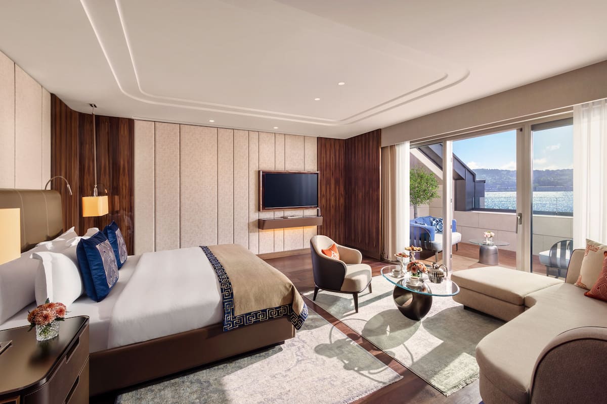 Quarto de hotel moderno com uma cama grande, TV, varanda com vista para a água e área de estar com mesa de vidro.