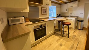 Uma cozinha moderna com armários brancos, forno embutido, micro-ondas e pia de aço inoxidável. Há uma janela que proporciona luz natural e teto de madeira.
