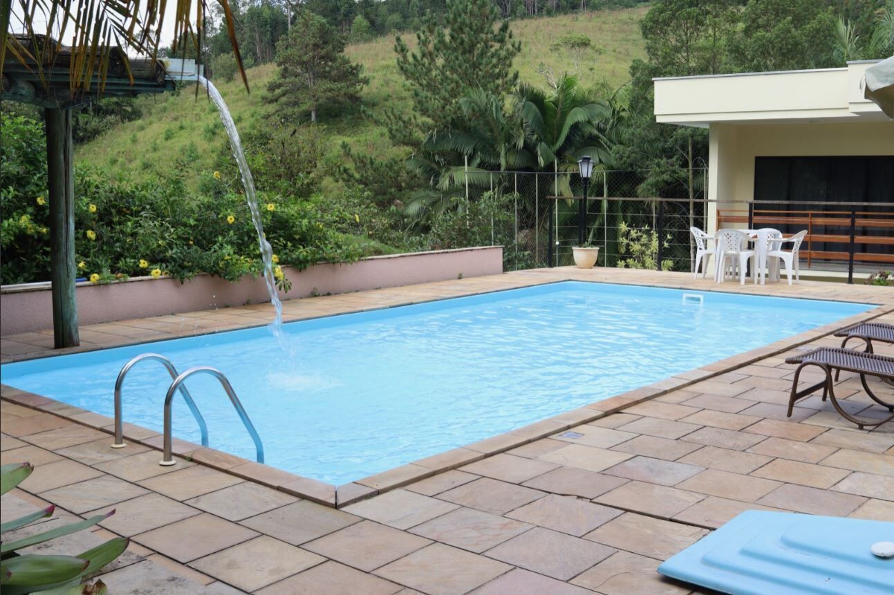 Piscina retangular com um recurso aquático, rodeada por um deck de azulejos, móveis de exterior e vista para uma paisagem acidentada.