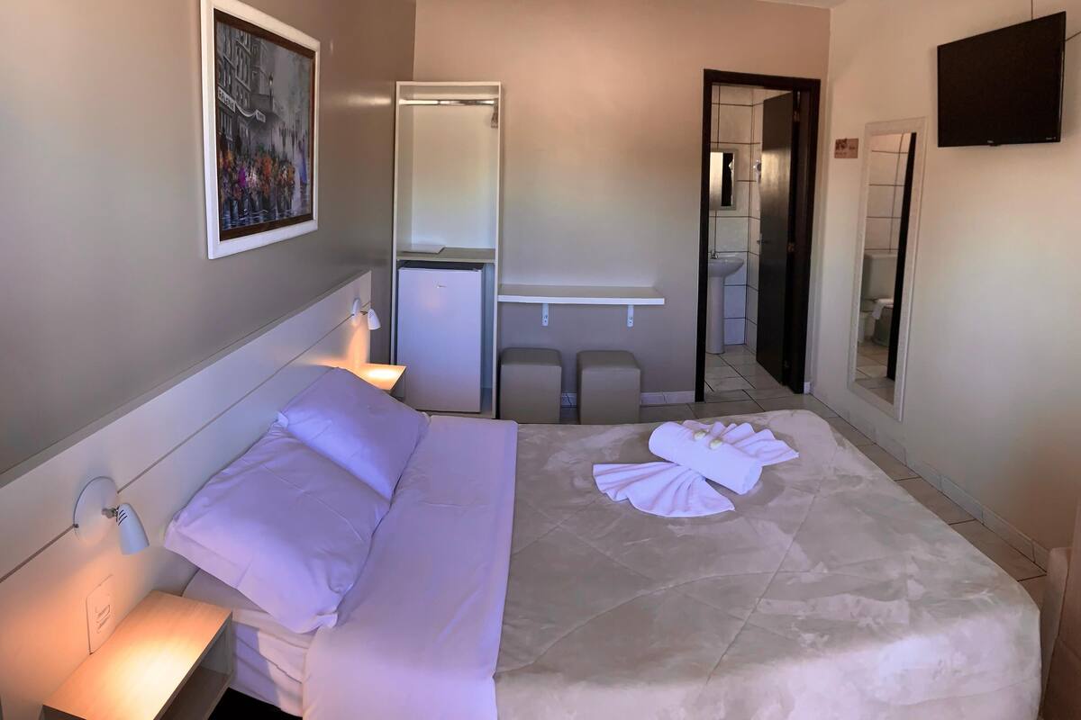 Quarto de hotel com cama, abajures, uma mesinha, espelho, TV e banheiro visível por uma porta aberta.