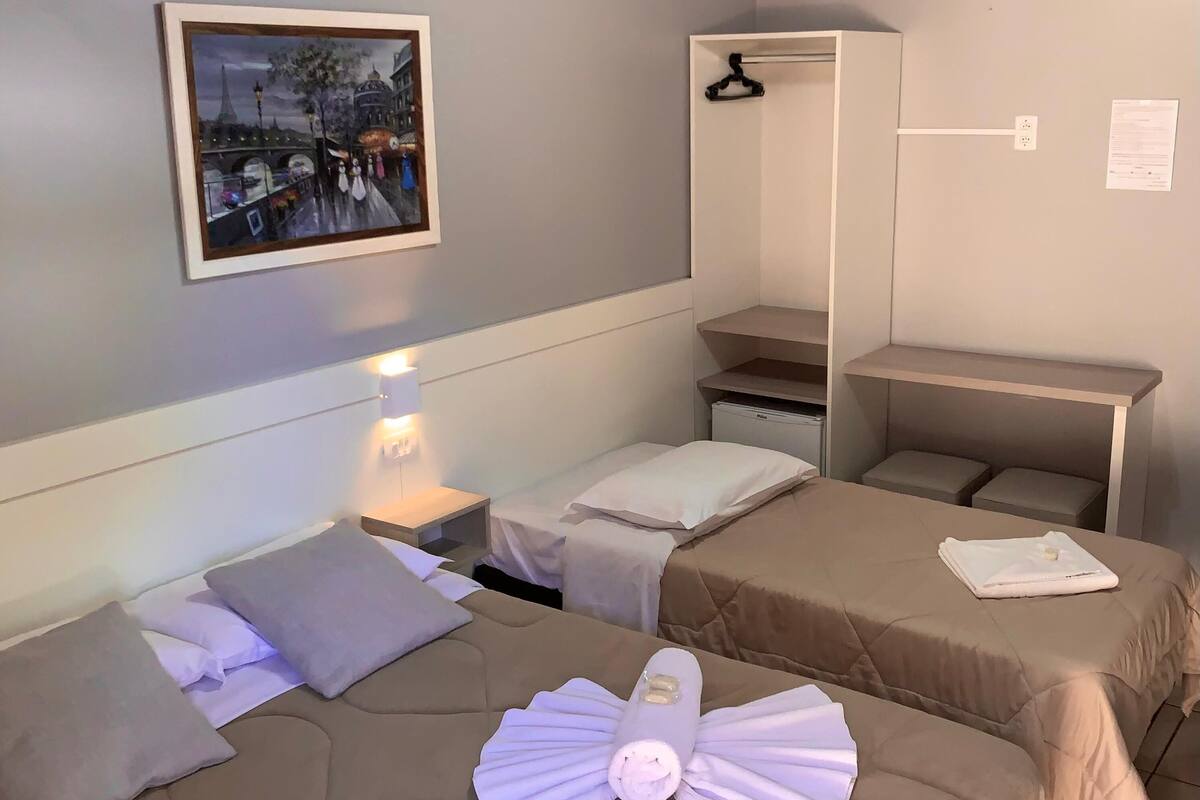 Quarto de hotel com duas camas, uma mesa de cabeceira, uma escrivaninha, um guarda-roupa e um quadro na parede.