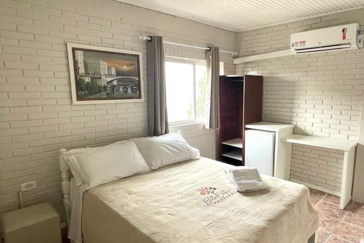 Quarto de hotel com uma cama, uma mesinha, um guarda-roupa e um aparelho de ar condicionado.