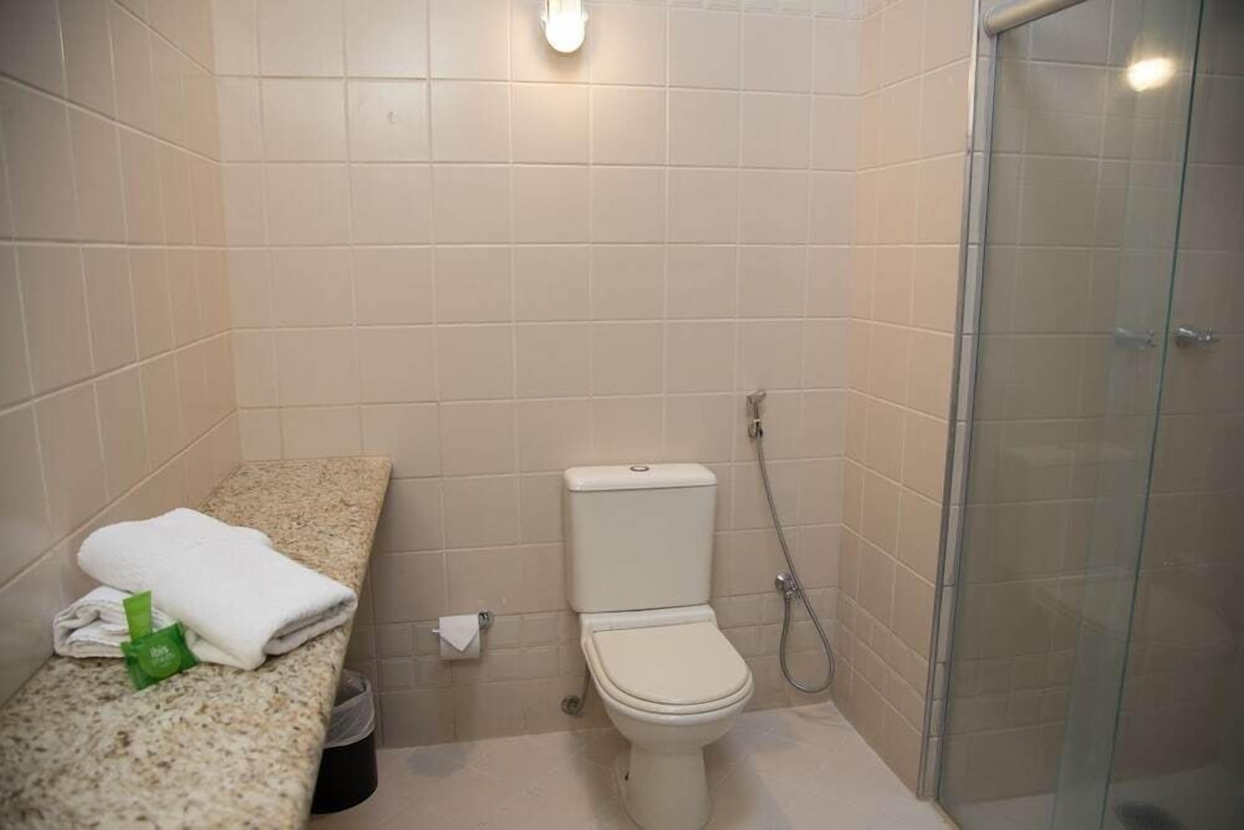 Casa de banho com sanita, cabine de duche em vidro e bancada com toalha e uma garrafa verde.