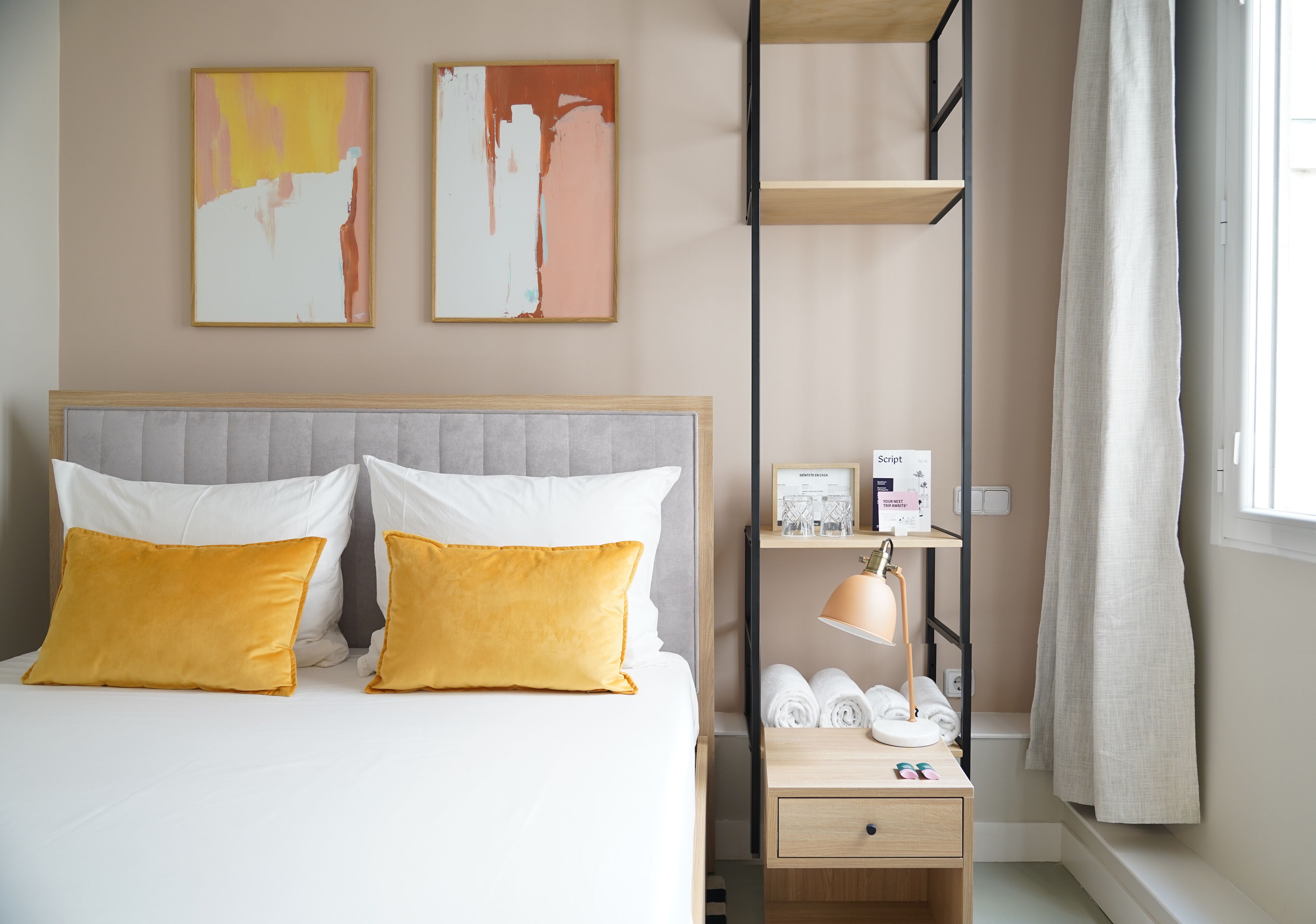 Quarto de hotel com uma cama, duas almofadas, uma mesa de cabeceira, uma luminária e uma prateleira com toalhas.