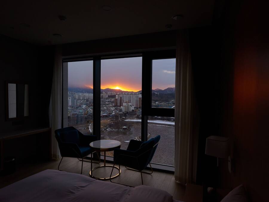 Quarto de hotel com uma janela grande, uma cama, duas cadeiras, uma mesa pequena e um abajur.