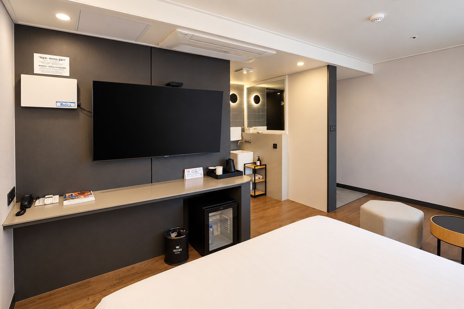 Quarto de hotel com cama grande, TV de tela plana na parede, escrivaninha pequena com telefone e revista, lixeira e geladeira compacta.