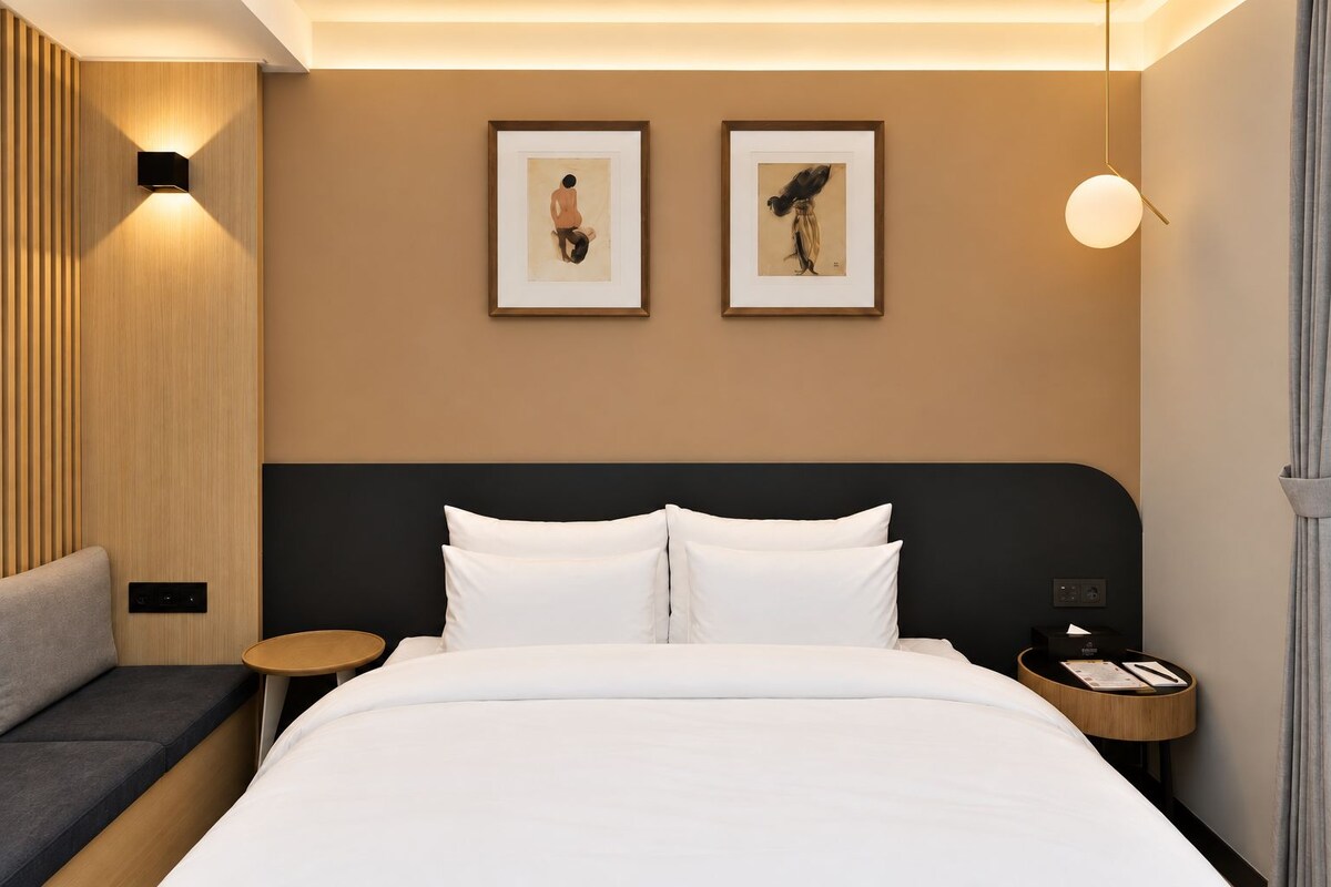 Quarto de hotel com cama, mesas de cabeceira, luminária na parede e duas obras de arte emolduradas.