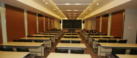 Sala de conferências com fileiras de mesas e cadeiras, equipada com um projetor na frente.