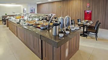 Uma área de buffet com diversos alimentos, incluindo frutas, doces e bebidas, com uma parede de painéis de madeira e um extintor fixado nela.