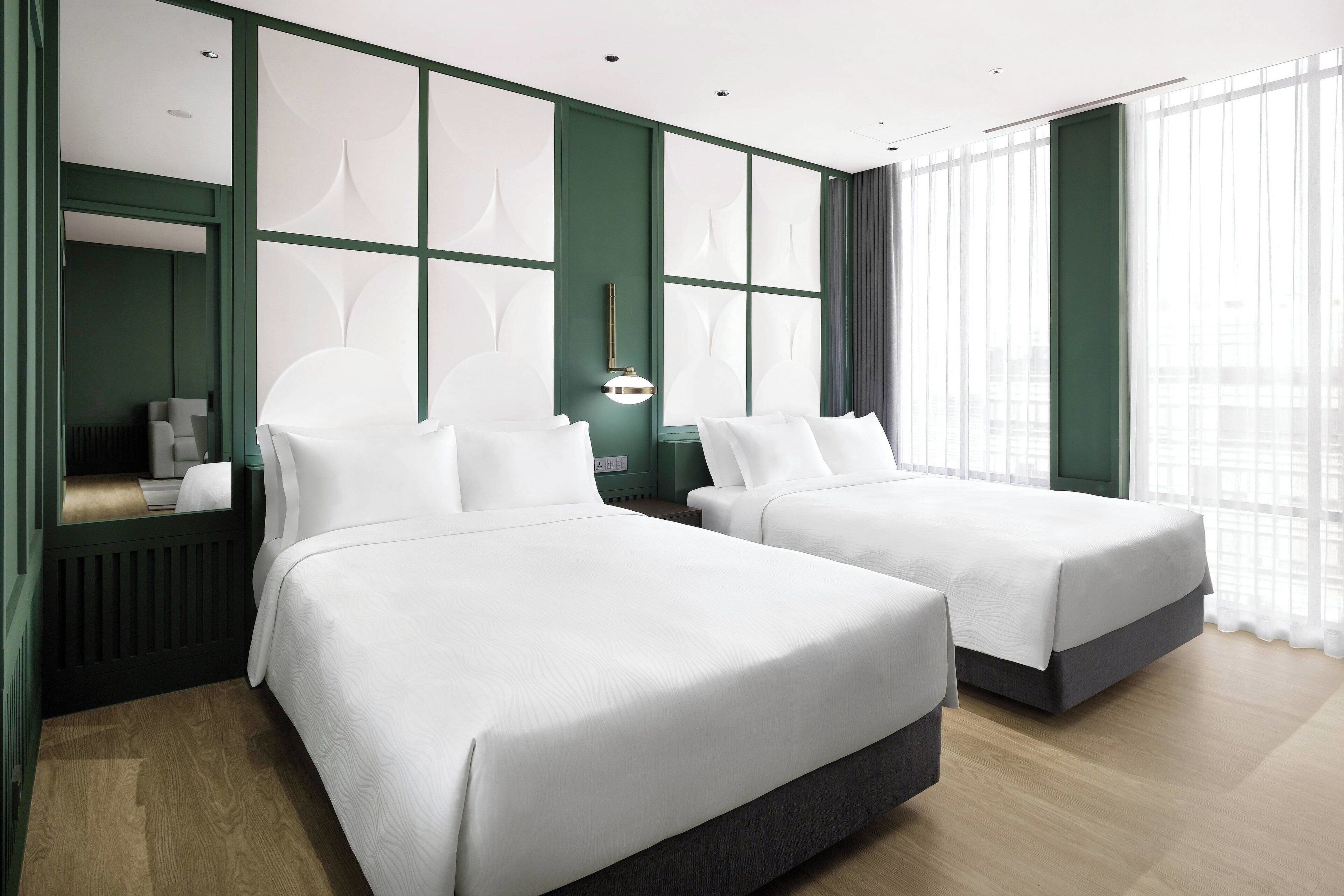 Quarto de hotel moderno com duas camas, uma mesa de trabalho e uma janela grande.