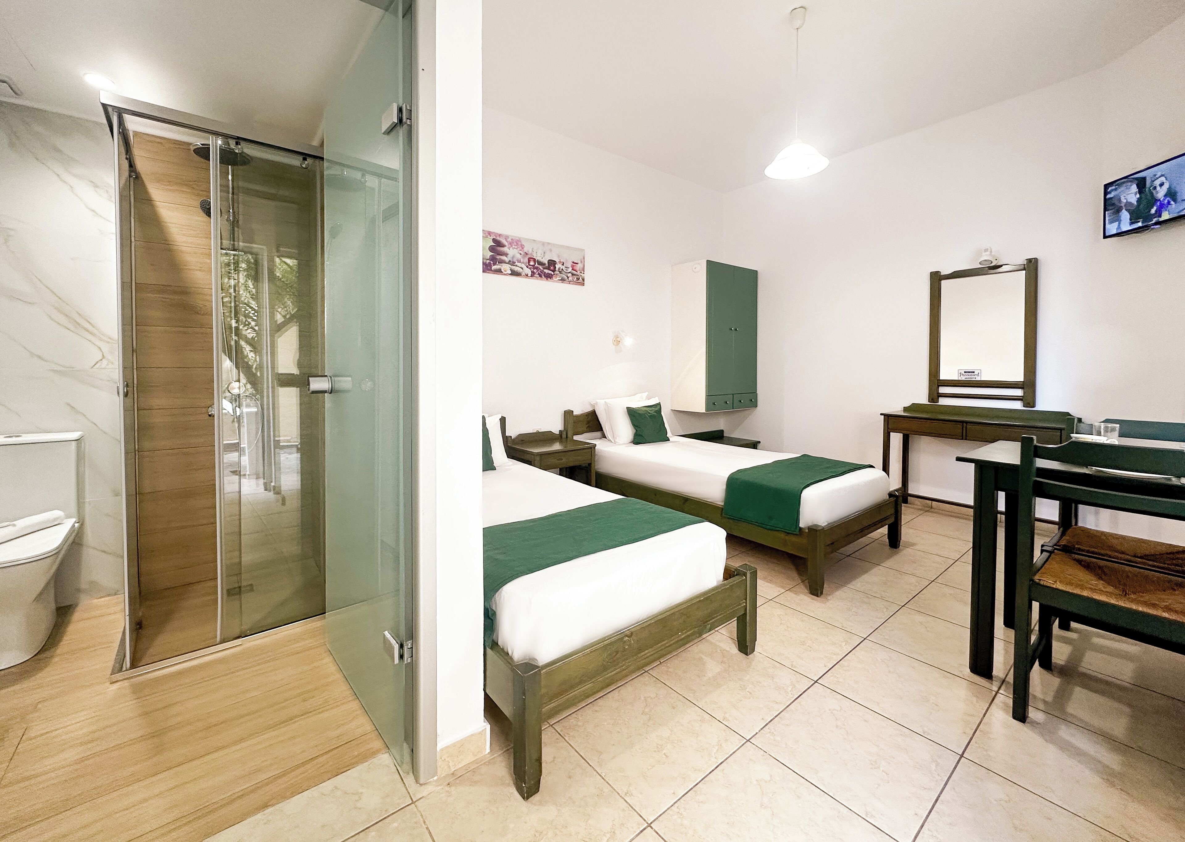 Quarto de hotel com chuveiro, duas camas, escrivaninha, cadeira, espelho e televisão.
