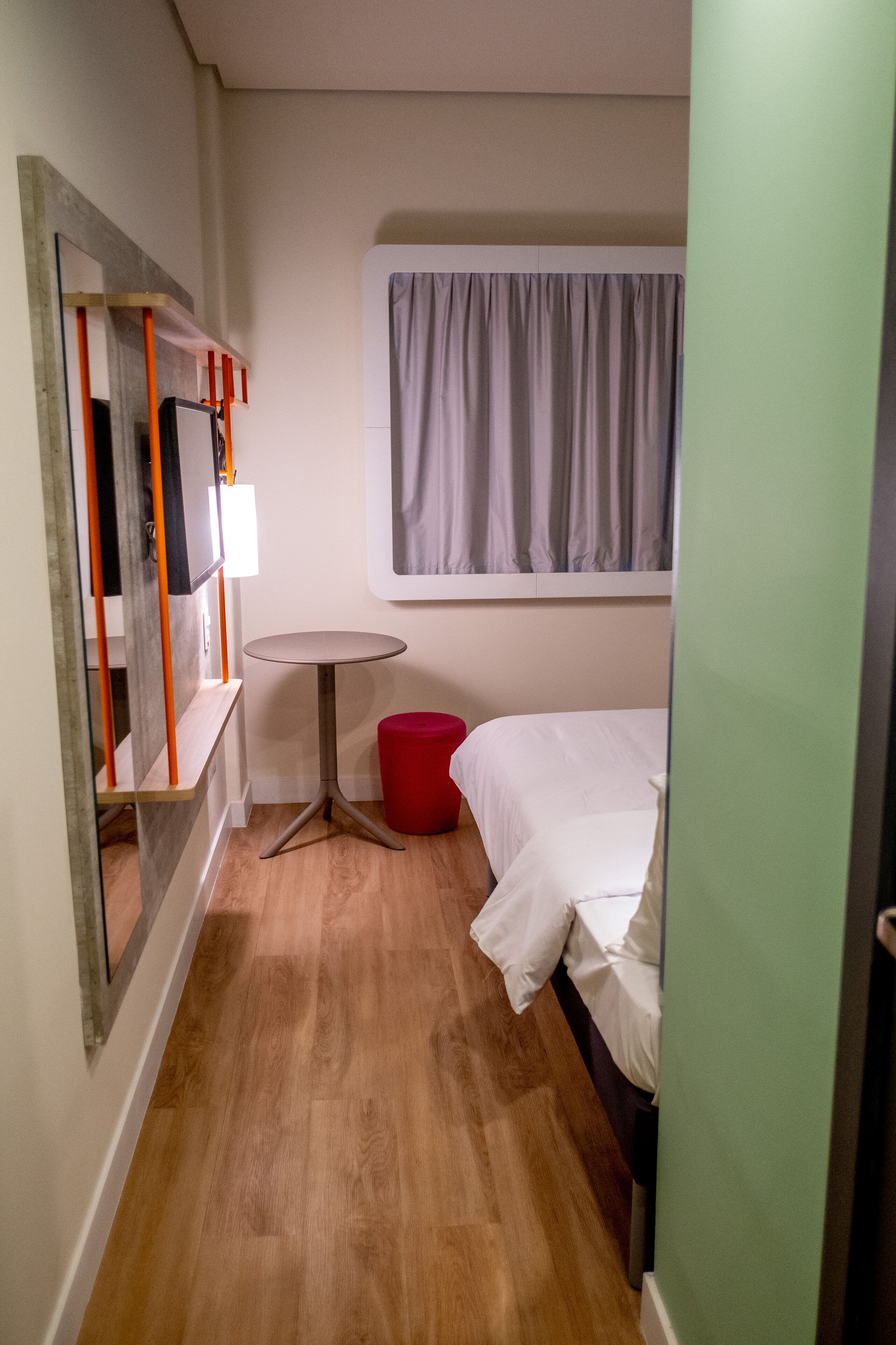 Quarto pequeno e aconchegante com uma cama, uma mesa redonda, um banquinho vermelho e uma janela com cortinas.